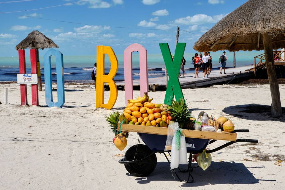 Uma praia com uma placa colorida que diz "NO BOX" e um carrinho de frutas.