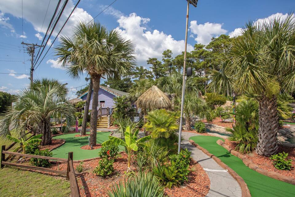 Um jardim tropical com palmeiras, uma construção de colmo e um campo de minigolfe.