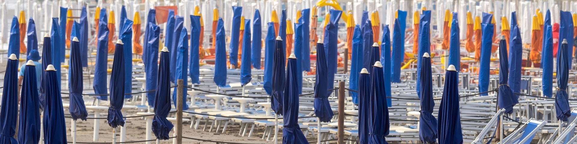 Fileiras de guarda-sóis e cadeiras de praia numa areia dourada, com um barco vermelho em primeiro plano.