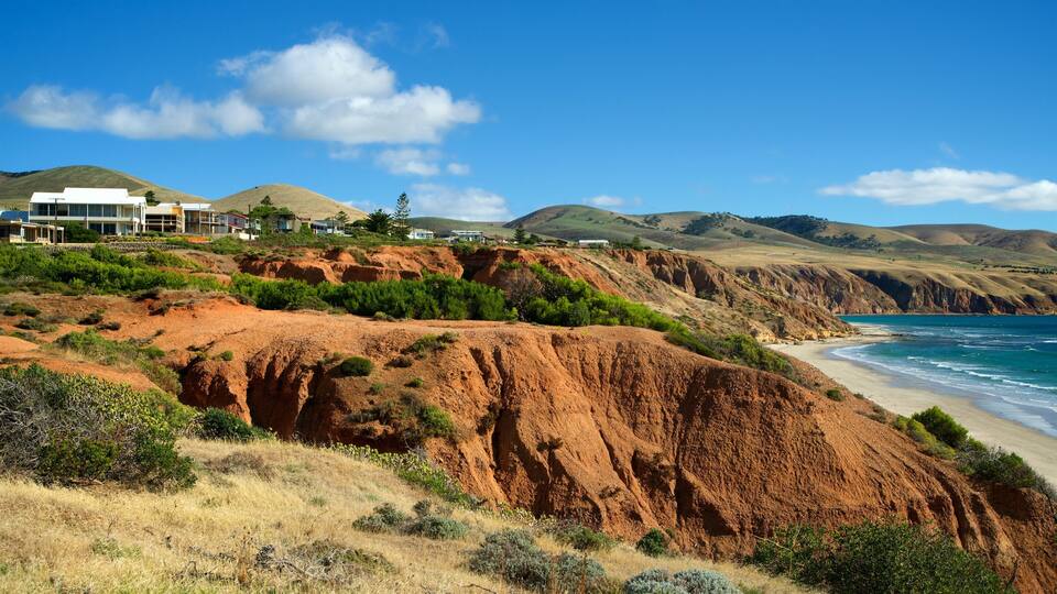 Paisagem costeira com falésias vermelhas, uma praia e casas na encosta.
