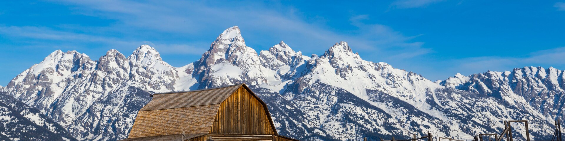 Um celeiro de madeira em uma paisagem nevada, com montanhas cobertas de neve ao fundo.