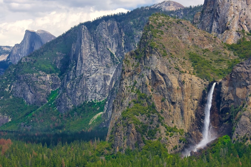 Uma cascata desce por um penhasco íngreme, rodeada por floresta densa e montanhas.