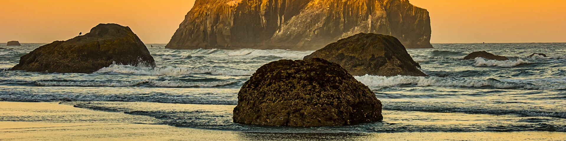 Costa rochosa com ondas ao pôr do sol, gaivotas na areia e uma formação rochosa imponente ao fundo.