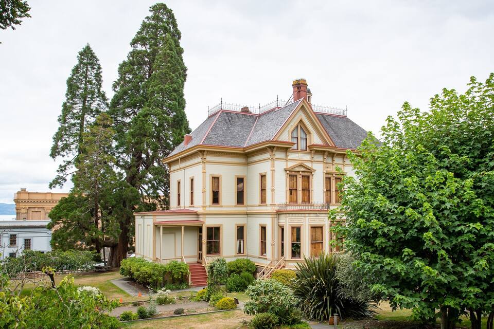 Uma casa grande e histórica com telhado vermelho e paredes brancas, cercada por vegetação exuberante e árvores altas.