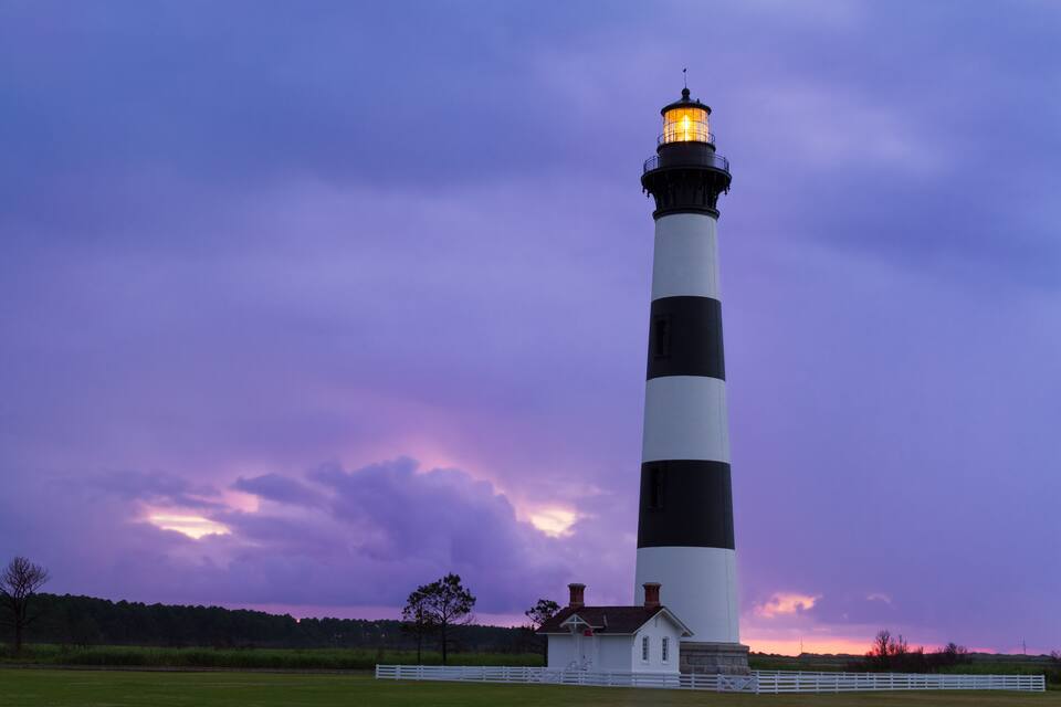 Um farol alto com listras pretas e brancas alternadas, um pequeno edifício branco em sua base, e um céu roxo com nuvens.