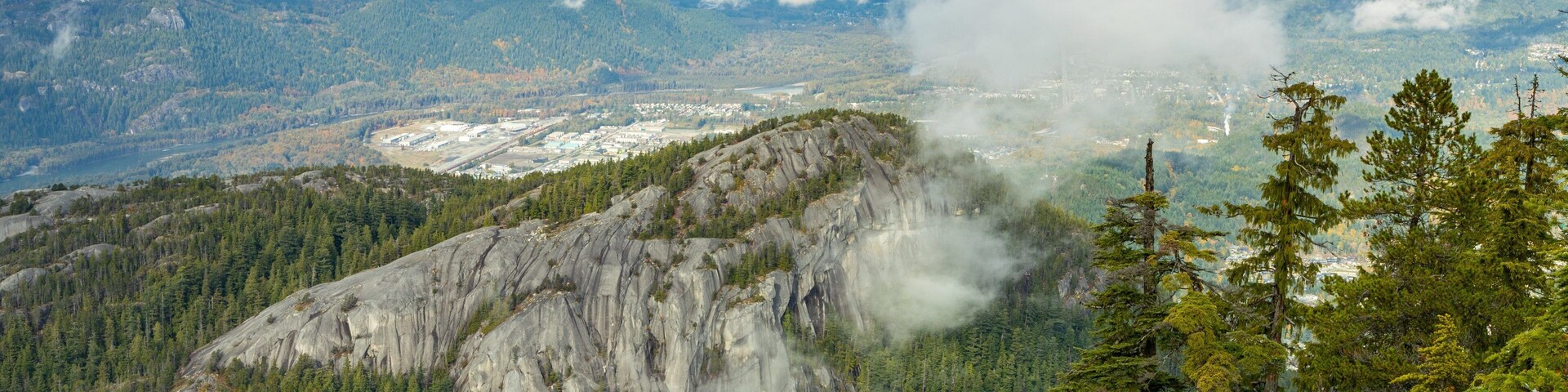 Paisagem montanhosa com florestas densas, afloramentos rochosos e uma vista distante de uma cidade.