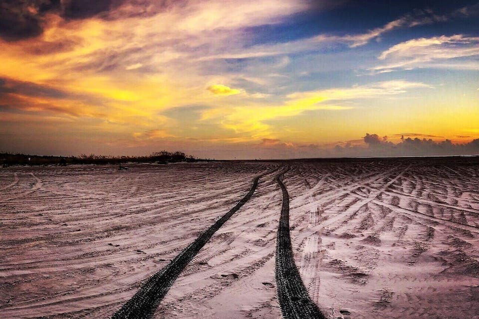 Paisagem nevada com marcas de pneus que se estendem até o horizonte sob um céu vibrante ao pôr do sol.