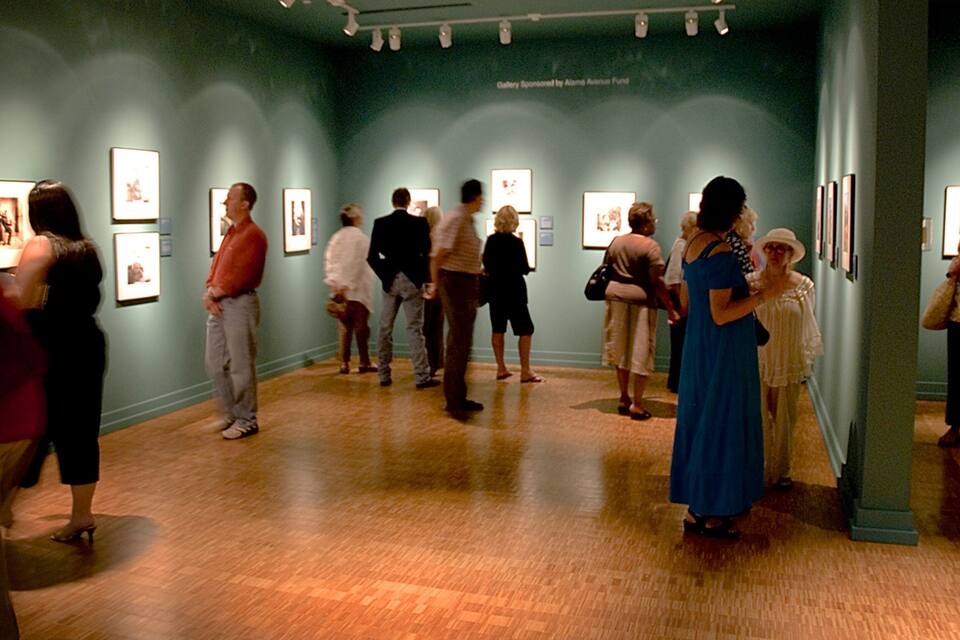 Uma galeria de arte com pessoas observando obras nas paredes.