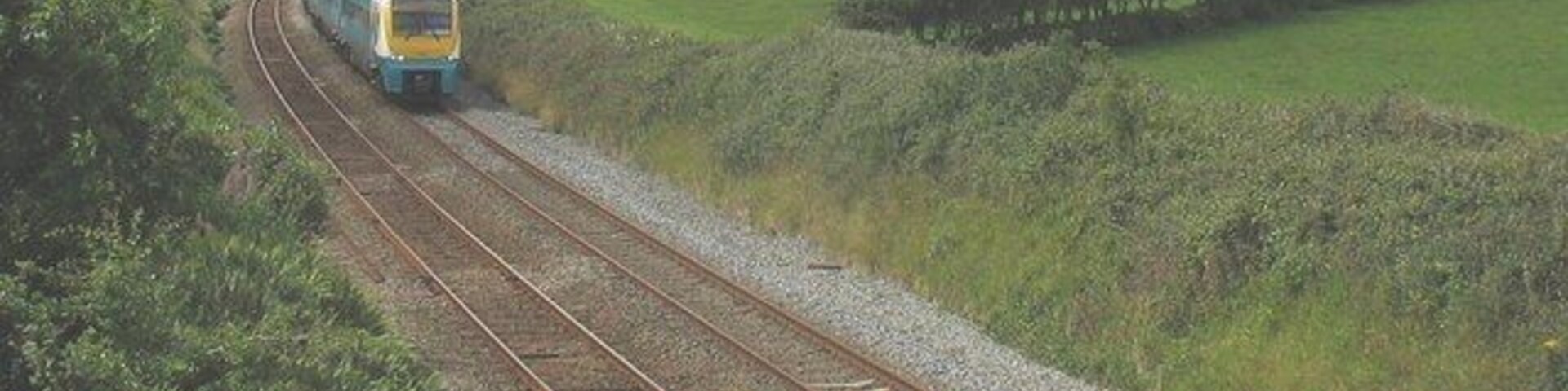 Um trem sobre trilhos em área rural, com campos verdes e árvores.