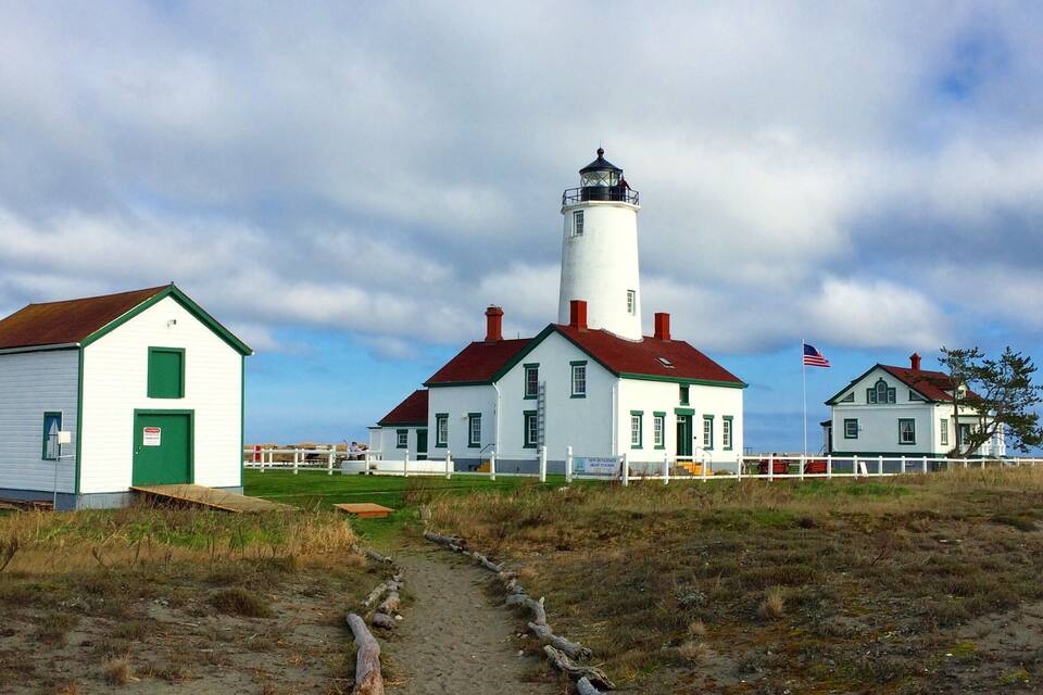 Um farol branco com teto vermelho, cercado por edifícios brancos com detalhes verdes, e um caminho de areia que leva até ele.
