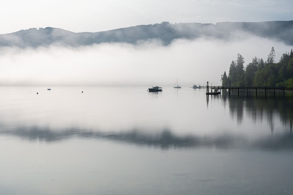 Um lago calmo com barcos, um cais e uma colina arborizada ao fundo.