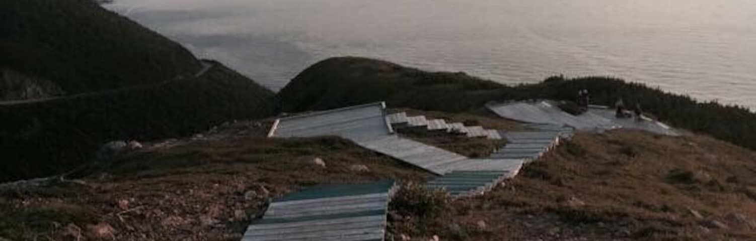 Um calçadão de madeira que leva a uma área costeira, com vista para o mar e colinas ao fundo.