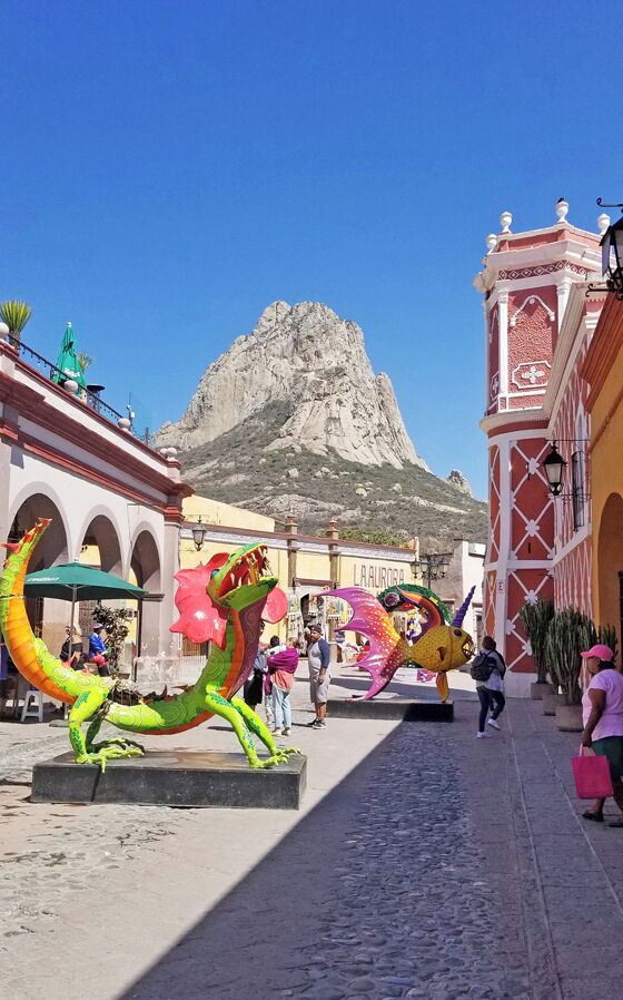 Uma praça com coloridas esculturas de dragões, uma montanha ao fundo e arquitetura tradicional.