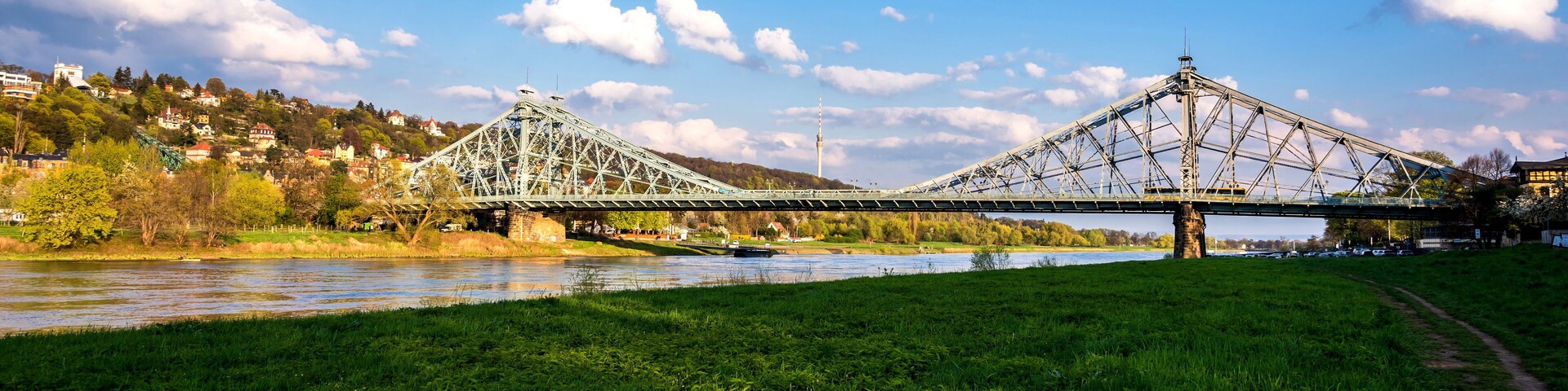 Uma grande ponte de aço atravessa um rio, com um horizonte urbano ao fundo.