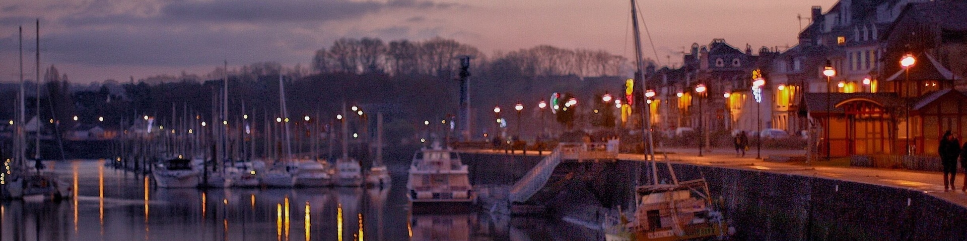 Um porto ao entardecer, com barcos atracados, uma ponte e edifícios iluminados.