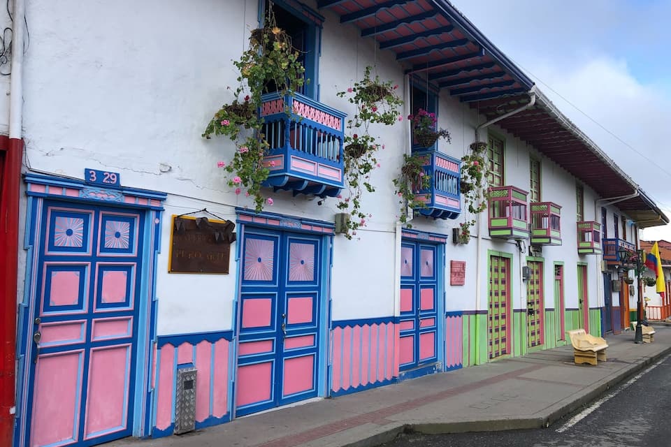 Um edifício colorido com portas azuis e cor-de-rosa, cestos de flores pendentes e uma rua com bancos.