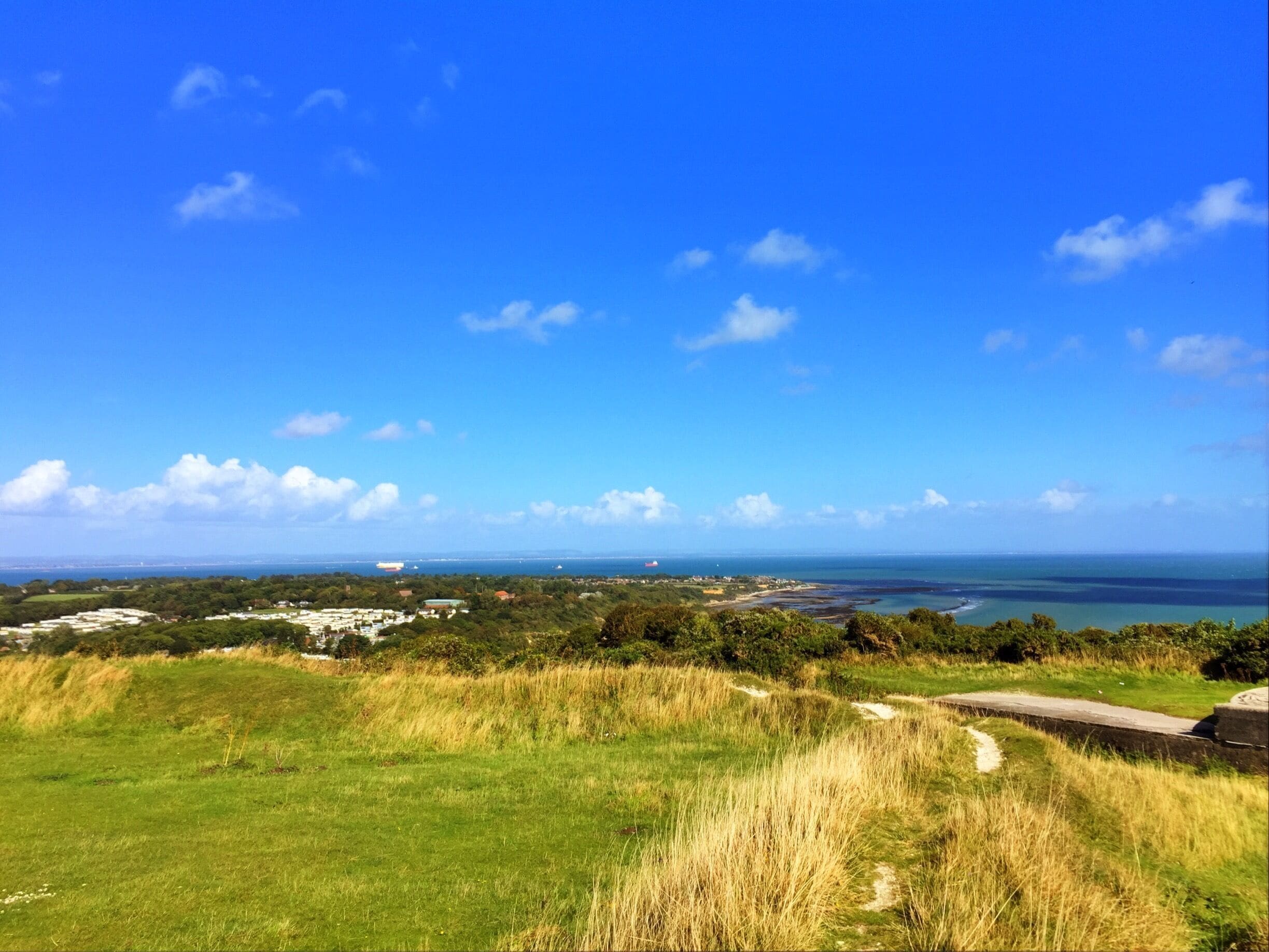 Paisagem costeira com céu azul límpido, campos verdes e vista distante do oceano.