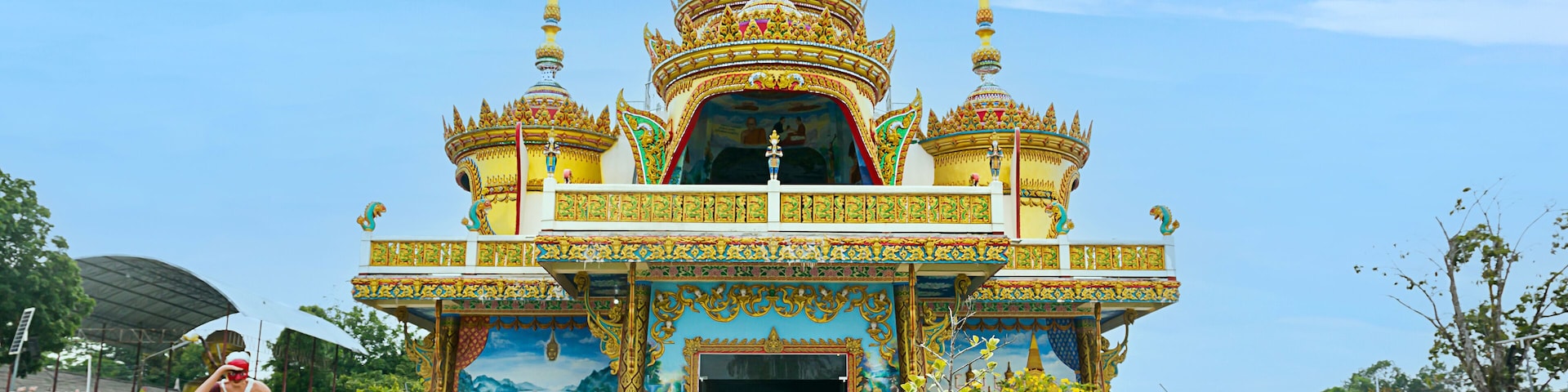 Um templo colorido com telhado dourado e várias torres, rodeado por vegetação exuberante e estátuas.