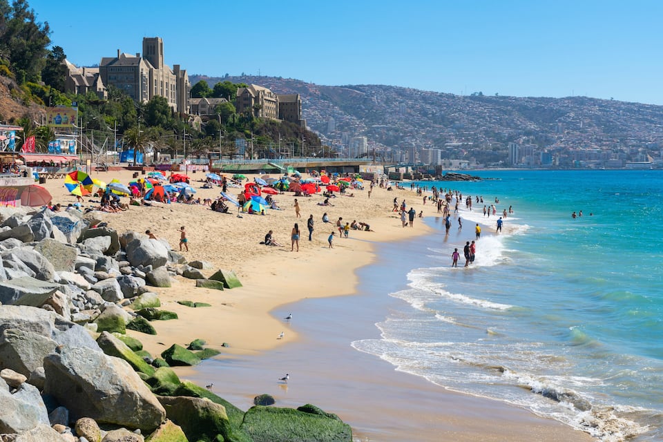 Uma praia movimentada com pessoas, guarda-sóis coloridos e pedras grandes.