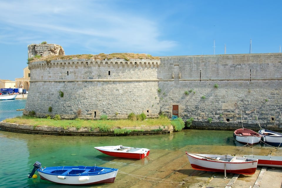 Uma fortificação de pedra junto a um porto calmo, com várias embarcações de pequeno porte atracadas.