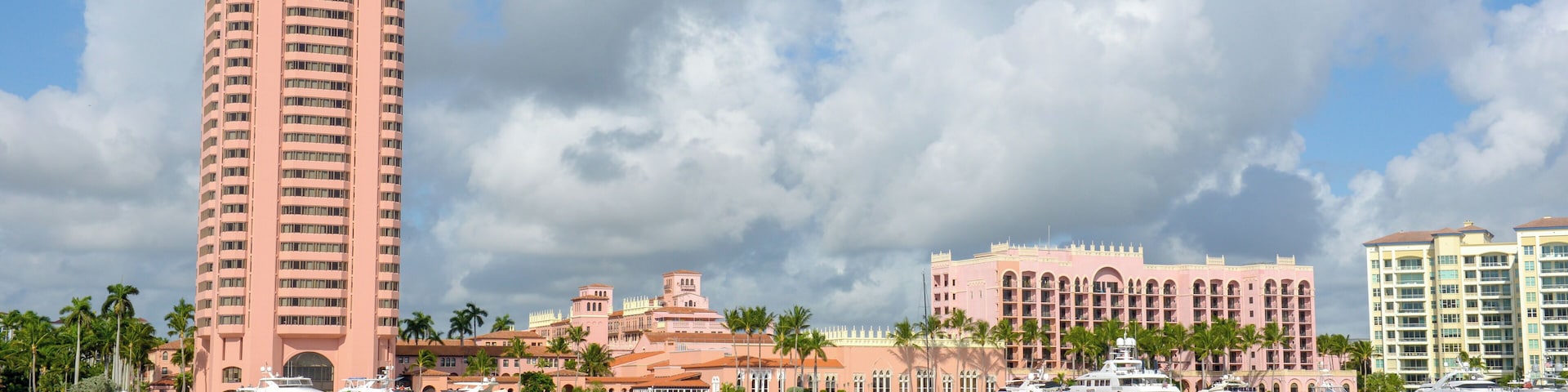 Uma orla com um edifício alto cor-de-rosa, vários edifícios menores cor-de-rosa e amarelos, e barcos atracados na costa.