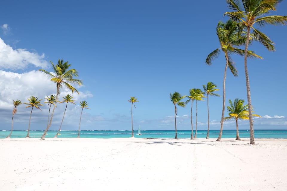 Uma praia com areia branca e águas azul-turquesa, palmeiras e um veleiro ao longe.