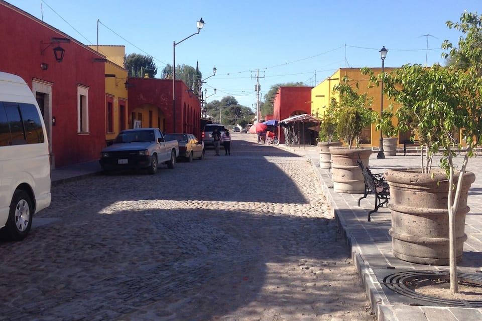 Ruas de paralelepípedos flanqueadas por edifícios coloridos, carros estacionados e alguns pedestres.