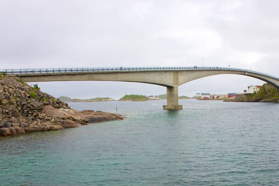 Uma ponte de concreto sobre um rio calmo, com margens rochosas e edifícios ao longe.