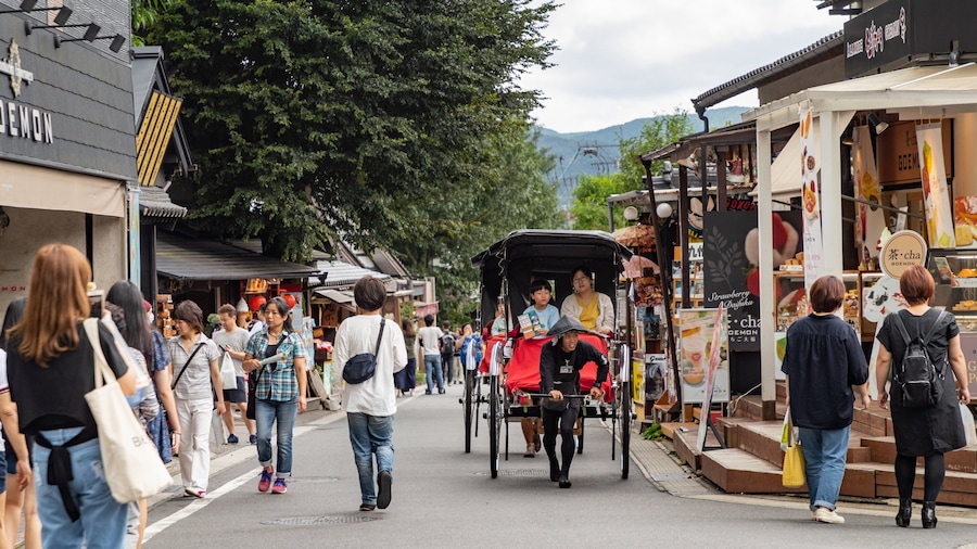Uma rua movimentada com pessoas caminhando, um rickshaw tradicional e diversas lojas.