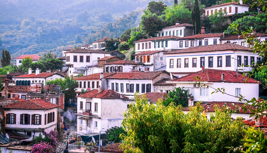 Uma vila em encosta com edifícios de telhados em terracota, vegetação exuberante e montanhas ao fundo.