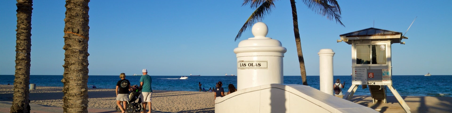 Uma praia com palmeiras, uma torre salva-vidas e pessoas caminhando.