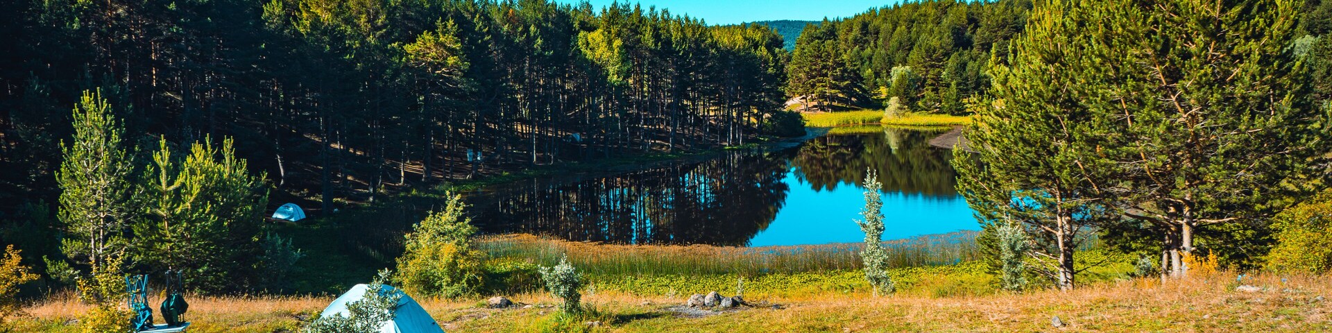 Um lago rodeado por uma floresta densa, uma tenda e um céu azul límpido.