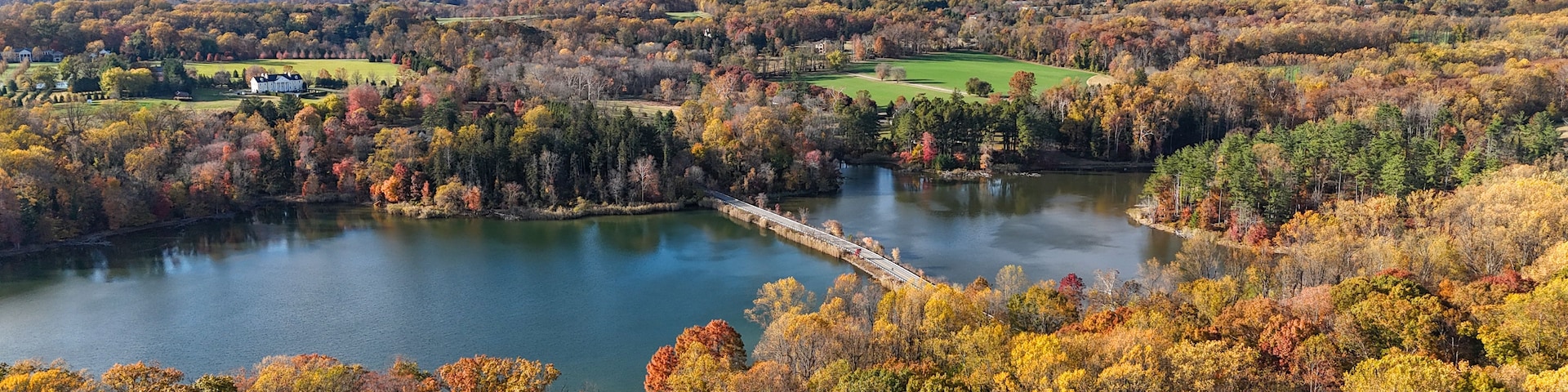 Um lago cercado por árvores com cores de outono, uma ponte e um campo de golfe ao longe.