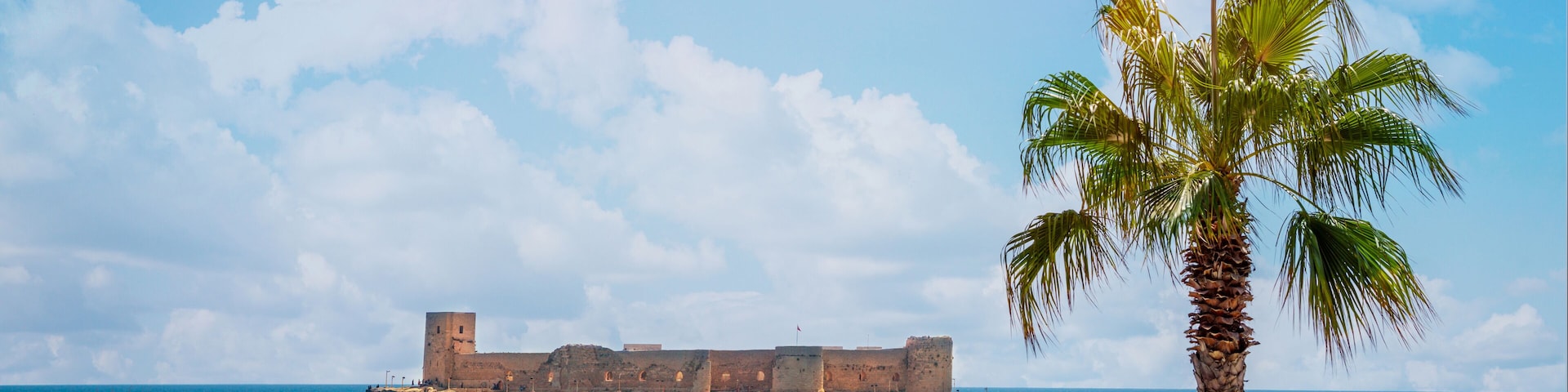 Uma paisagem costeira com um forte histórico, mar azul calmo e palmeiras.