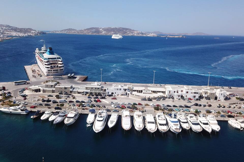 Uma marina com vários iates atracados, um grande cruzeiro e uma cidade costeira com edifícios brancos.