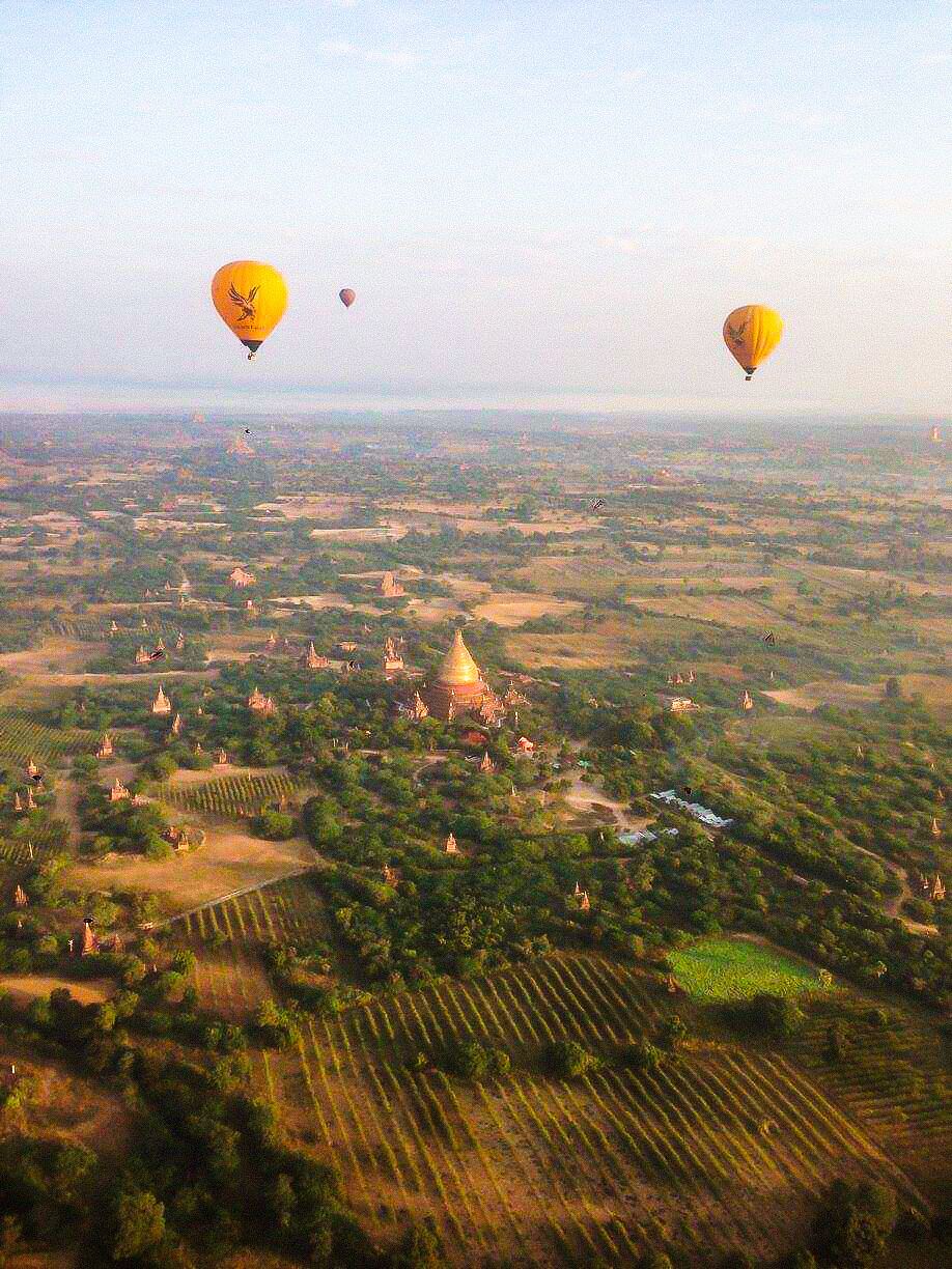 Balões de ar quente sobrevoando uma paisagem com campos, árvores e um templo.