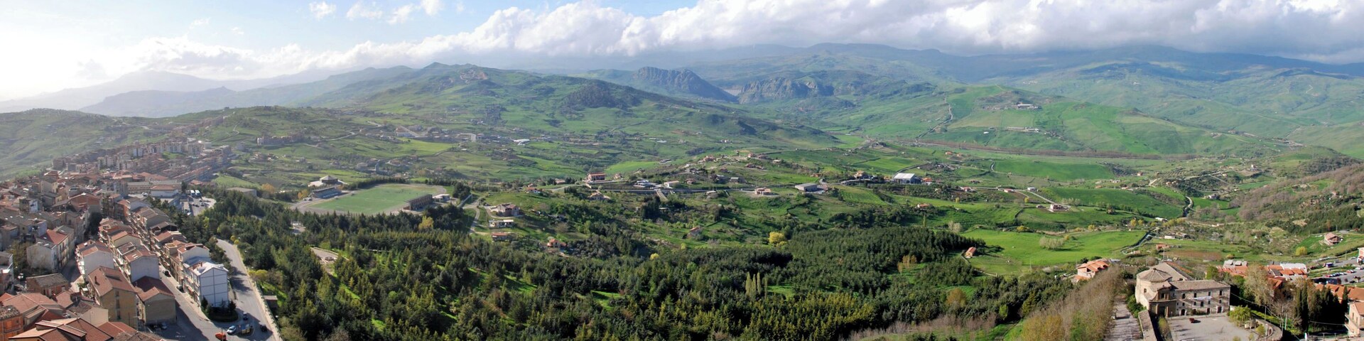 Vista panorâmica de uma cidade aninhada num vale, com colinas ondulantes e uma cadeia de montanhas ao longe.