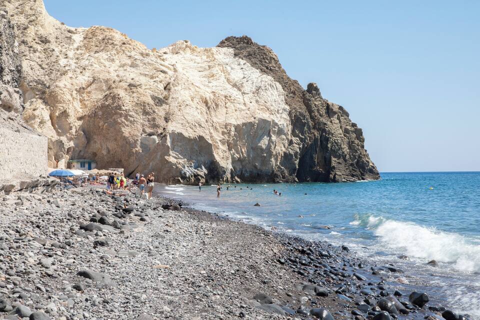 Uma praia rochosa com pessoas nadando e um grande penhasco ao fundo.