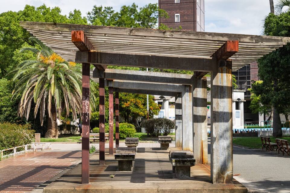 Uma pérgula de madeira com bancos num parque, com árvores e um edifício ao fundo.