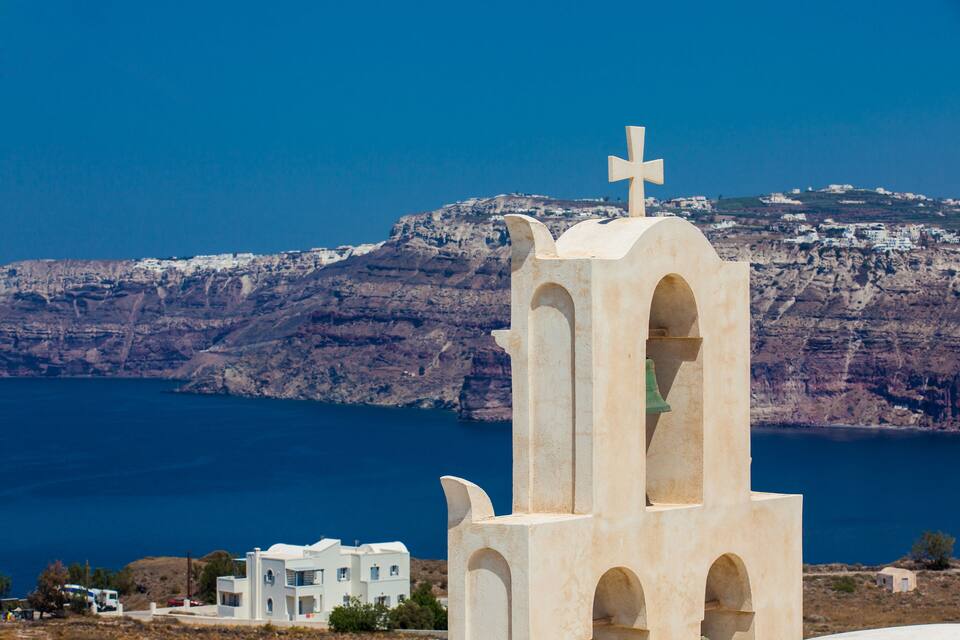 Igreja branca com cruz no topo, com vista para o mar azul e falésias rochosas.