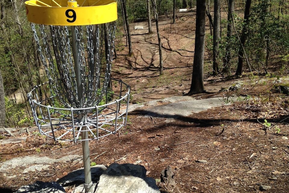 Cesto de disc golf identificado com o número 9 numa área arborizada, com um caminho de terra e pedras espalhadas.