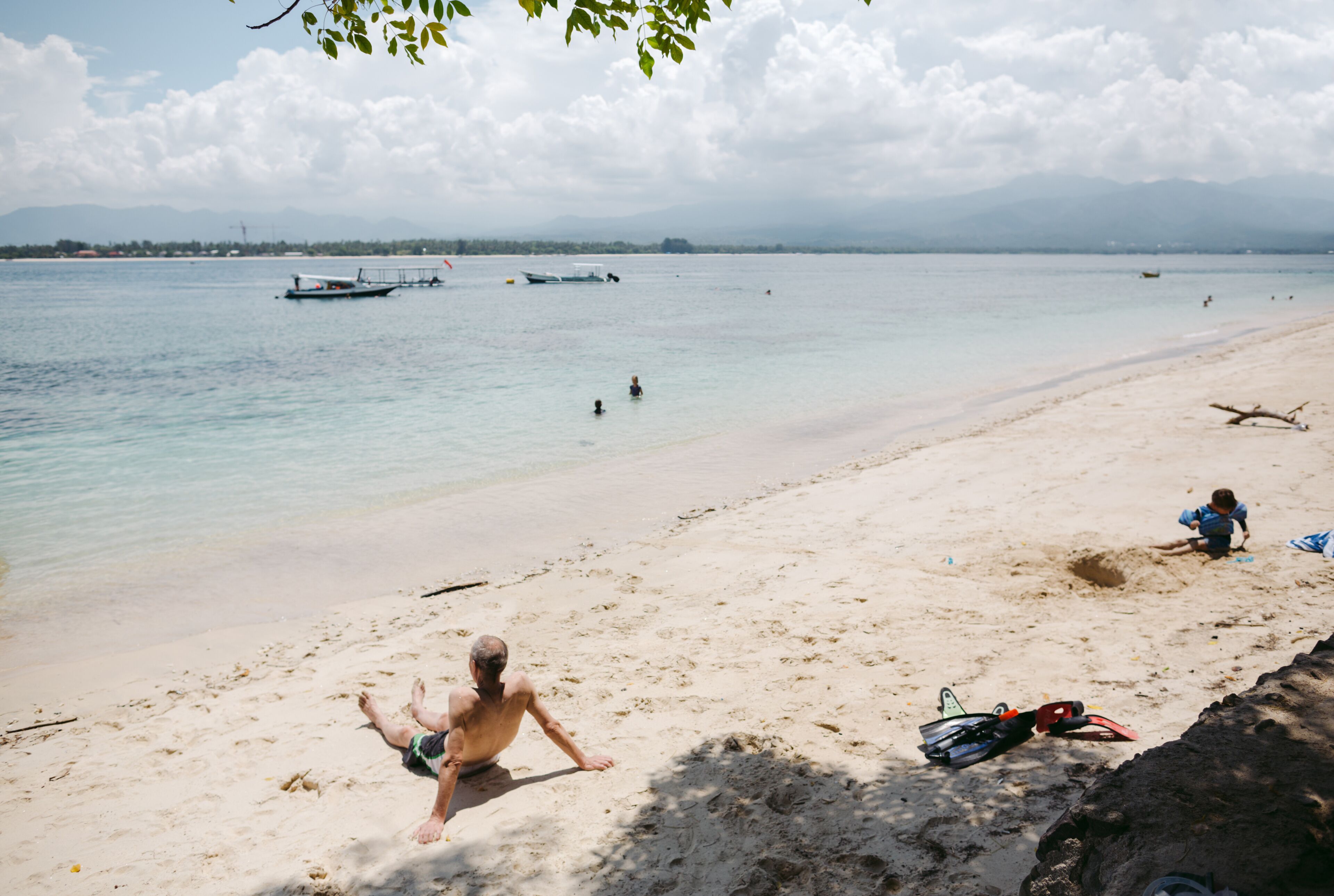 Praia arenosa com céu claro, mar calmo, barcos e pessoas.