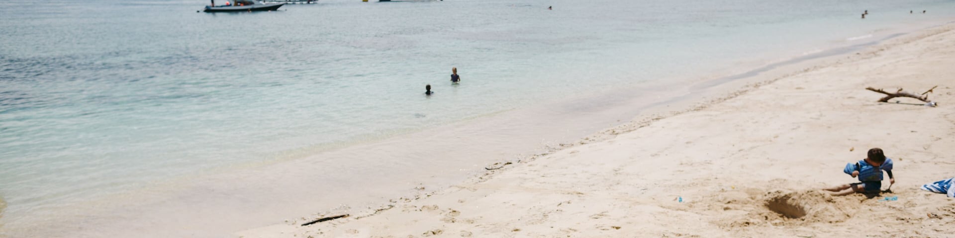 Praia arenosa com céu claro, mar calmo, barcos e pessoas.
