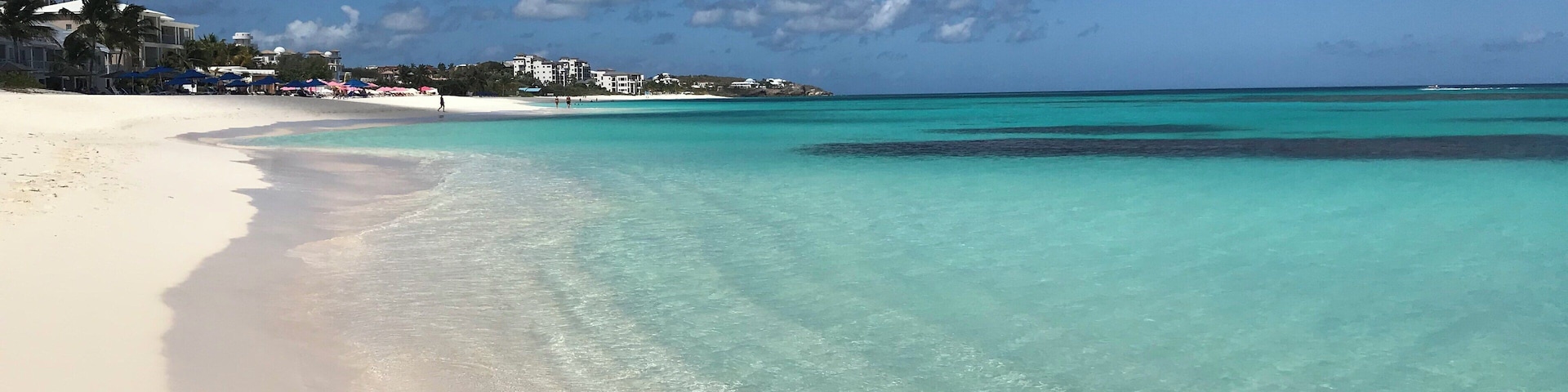 Uma praia com águas turquesa cristalinas, areia branca e edifícios ao fundo.