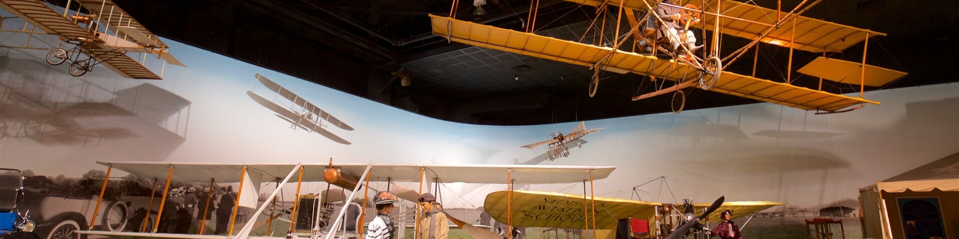 Exposição de museu com diversos biplanos históricos e um carro antigo.