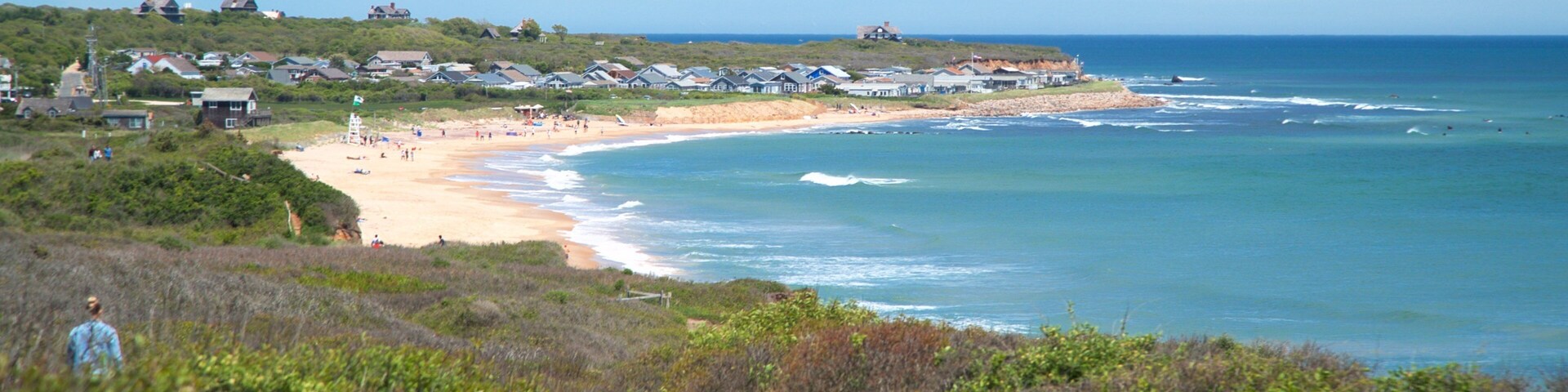 Paisagem costeira com praia de areia, ondas do oceano e casas numa encosta.