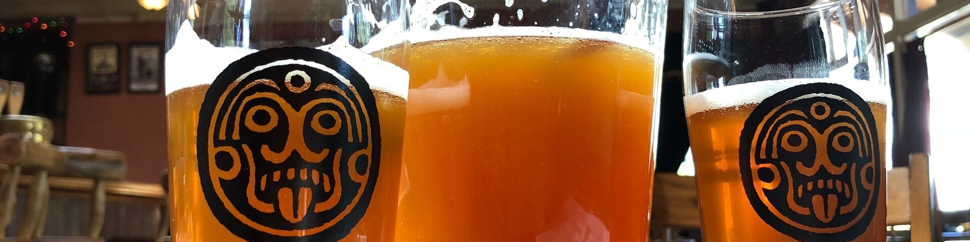 Três copos de cerveja com o logo de caveira, um copo alto e dois copos baixos, sobre uma mesa de madeira.