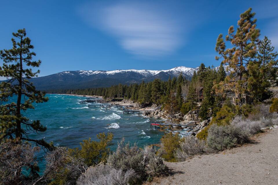 Paisagem costeira com céu azul límpido, costa rochosa e montanhas nevadas ao fundo.