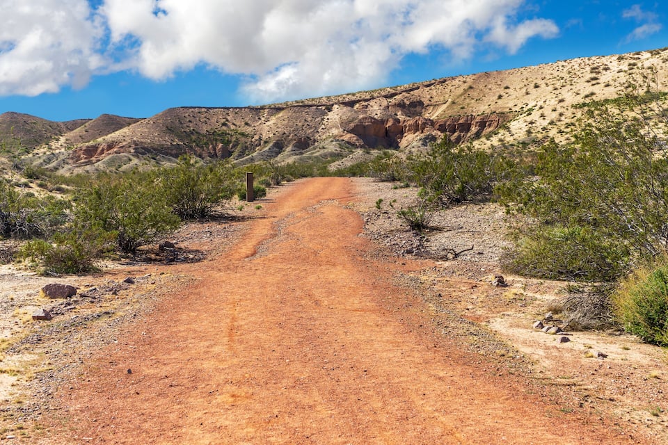 Uma estrada de terra serpenteia por uma paisagem desértica, com vegetação rala e colinas rochosas.