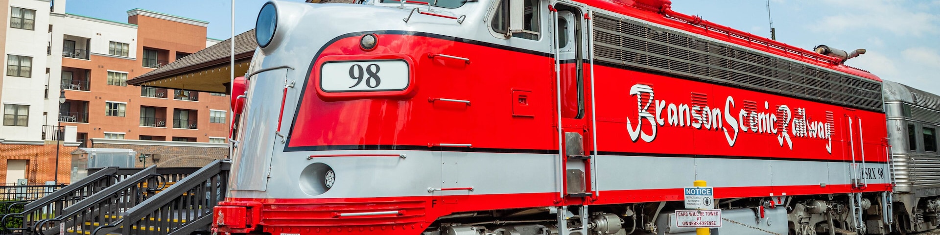 Um comboio Branson Scenic Railway vermelho e branco, com o número 98, estacionado nos carris com um horizonte urbano ao fundo.